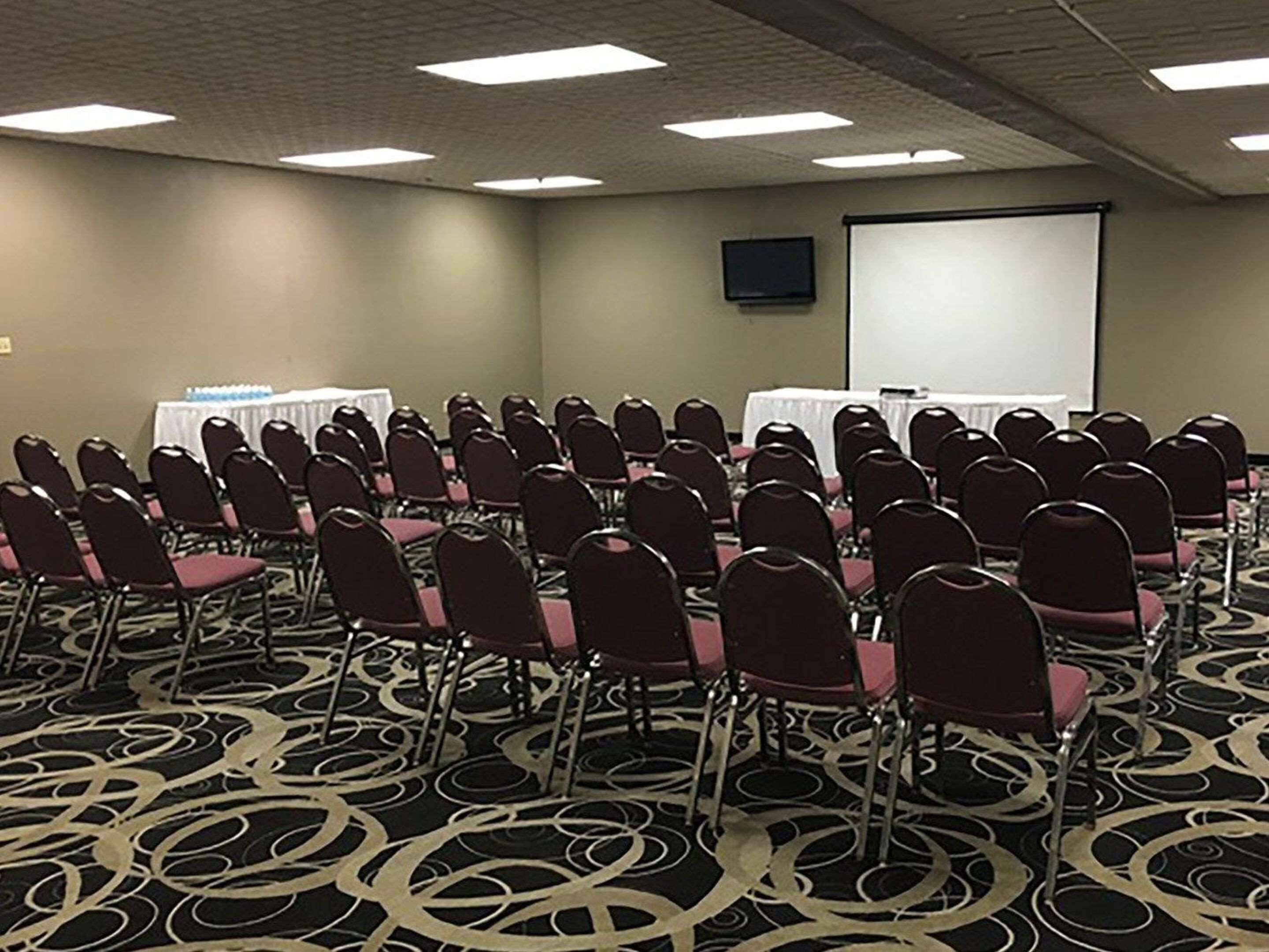Uma sala de conferências com fileiras de cadeiras, uma tela de projetor e uma mesa com garrafas de água.