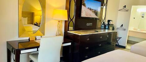 Quarto de hotel com cama, escrivaninha, cadeira, televisão, cômoda e espelho.