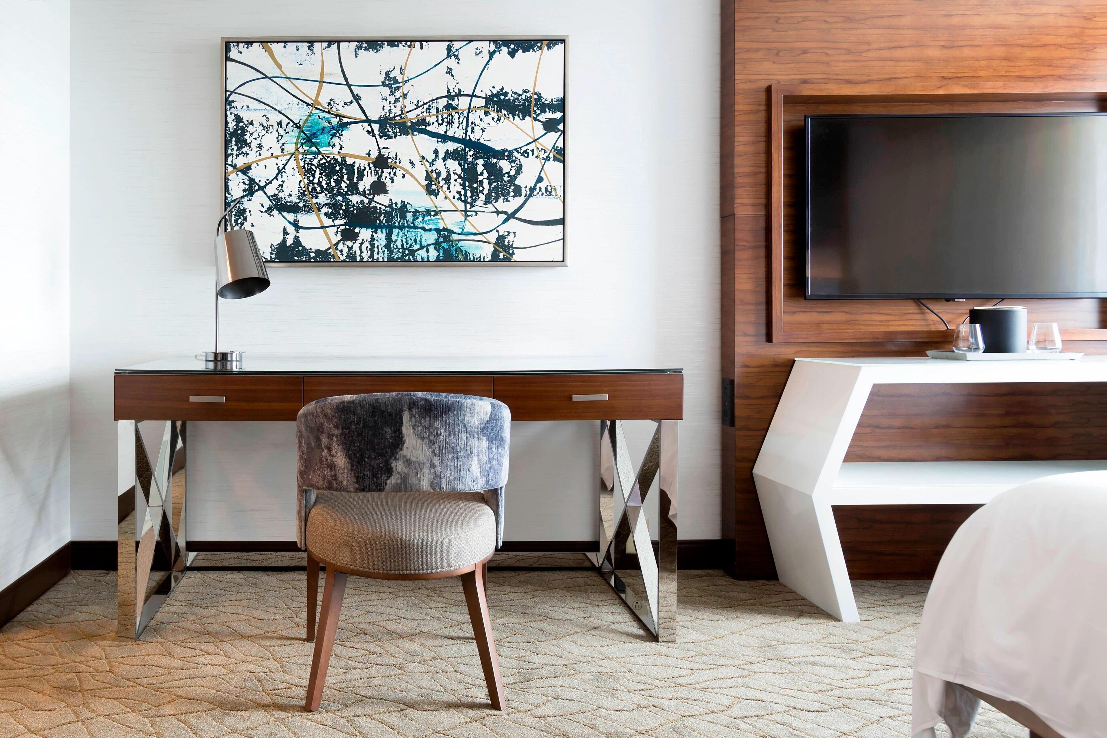 Quarto de hotel com uma escrivaninha moderna, uma cadeira, televisão de tela plana e um grande quadro abstrato na parede.