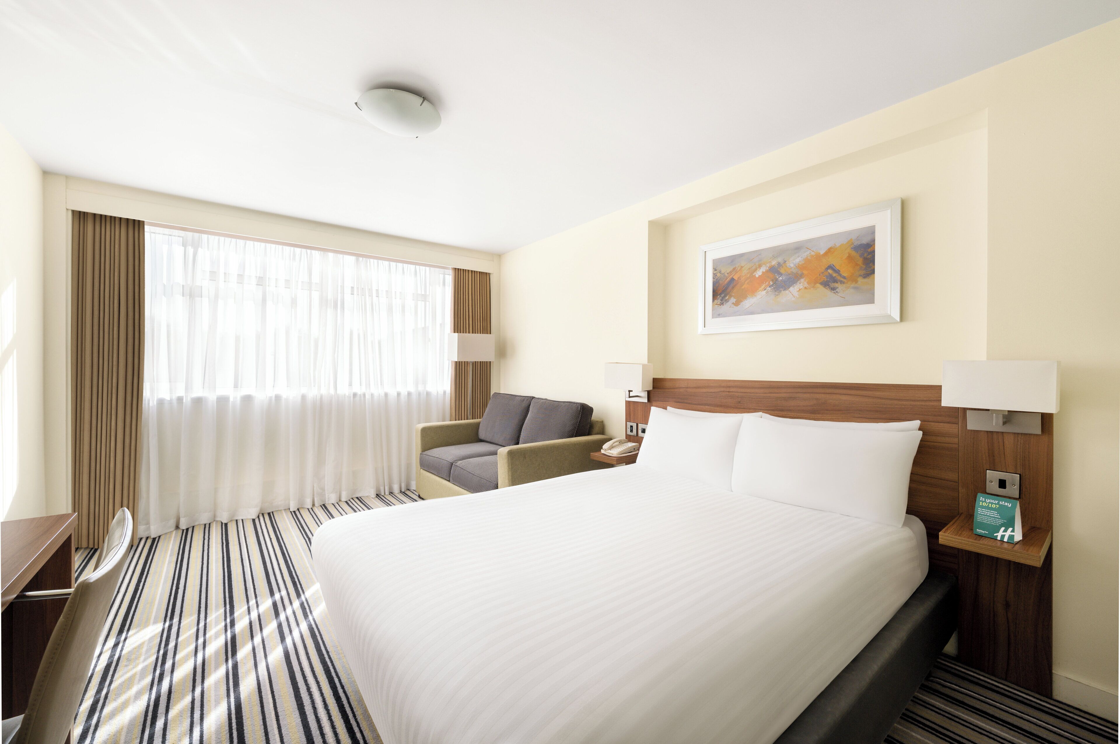 Quarto de hotel com uma cama grande, uma escrivaninha, uma cadeira, uma mesa de cabeceira e um quadro na parede.