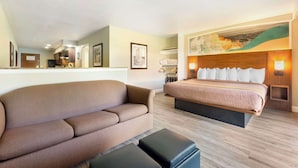 Quarto de hotel com sofá, cama com almofadas brancas, cabeceira de madeira e uma pintura grande na parede.