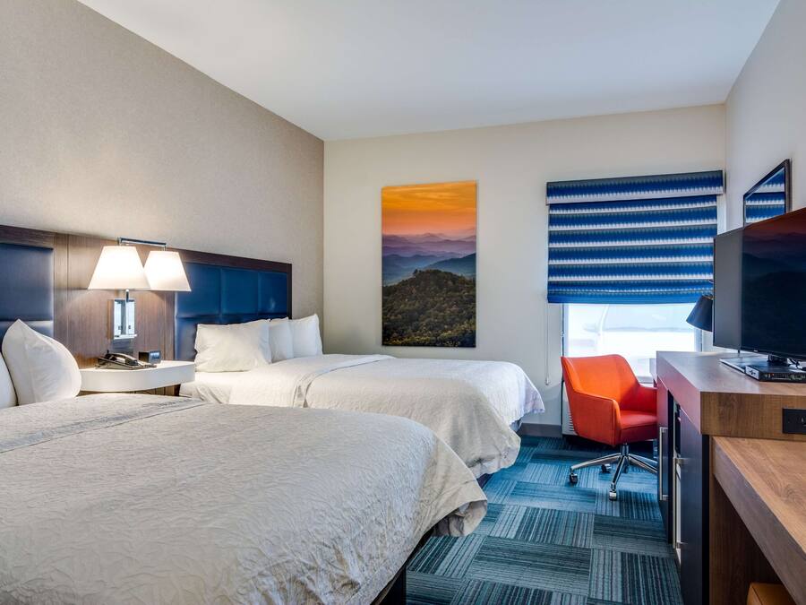 Quarto de hotel com duas camas, uma escrivaninha com televisão, uma cadeira e um quadro na parede.