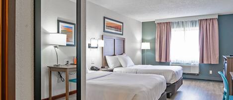 Quarto de hotel com duas camas, uma escrivaninha com luminária, uma cadeira e uma janela com cortinas.