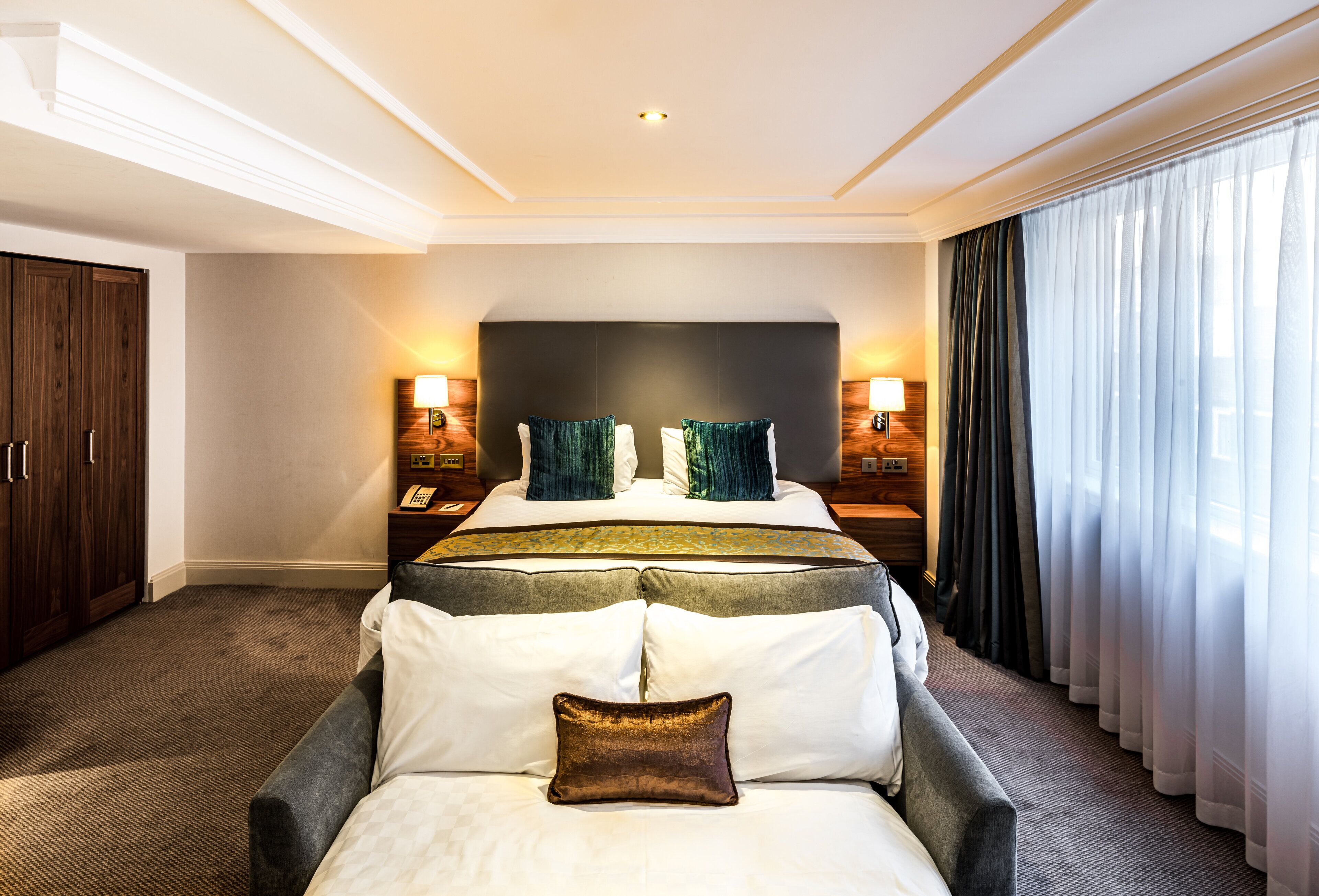 Quarto de hotel com uma cama grande, mesa de cabeceira, guarda-roupa e janela com cortinas.