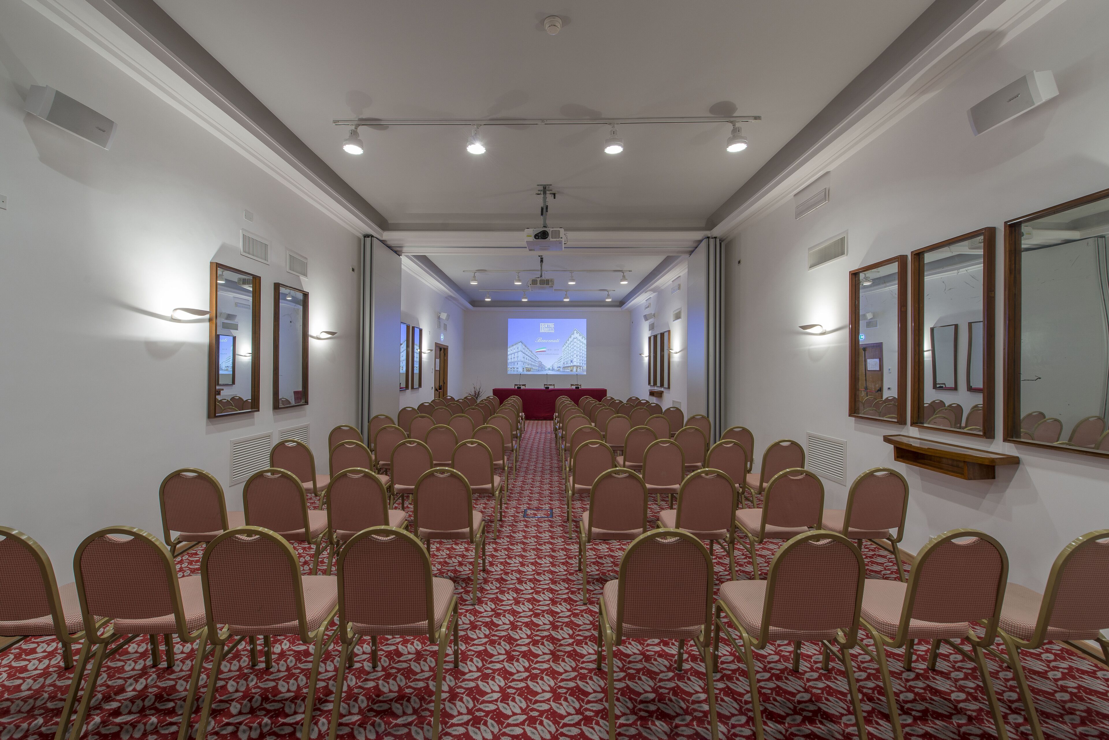 Sala de conferência com fileiras de cadeiras, piso coberto de carpete e uma tela de projeção na frente.