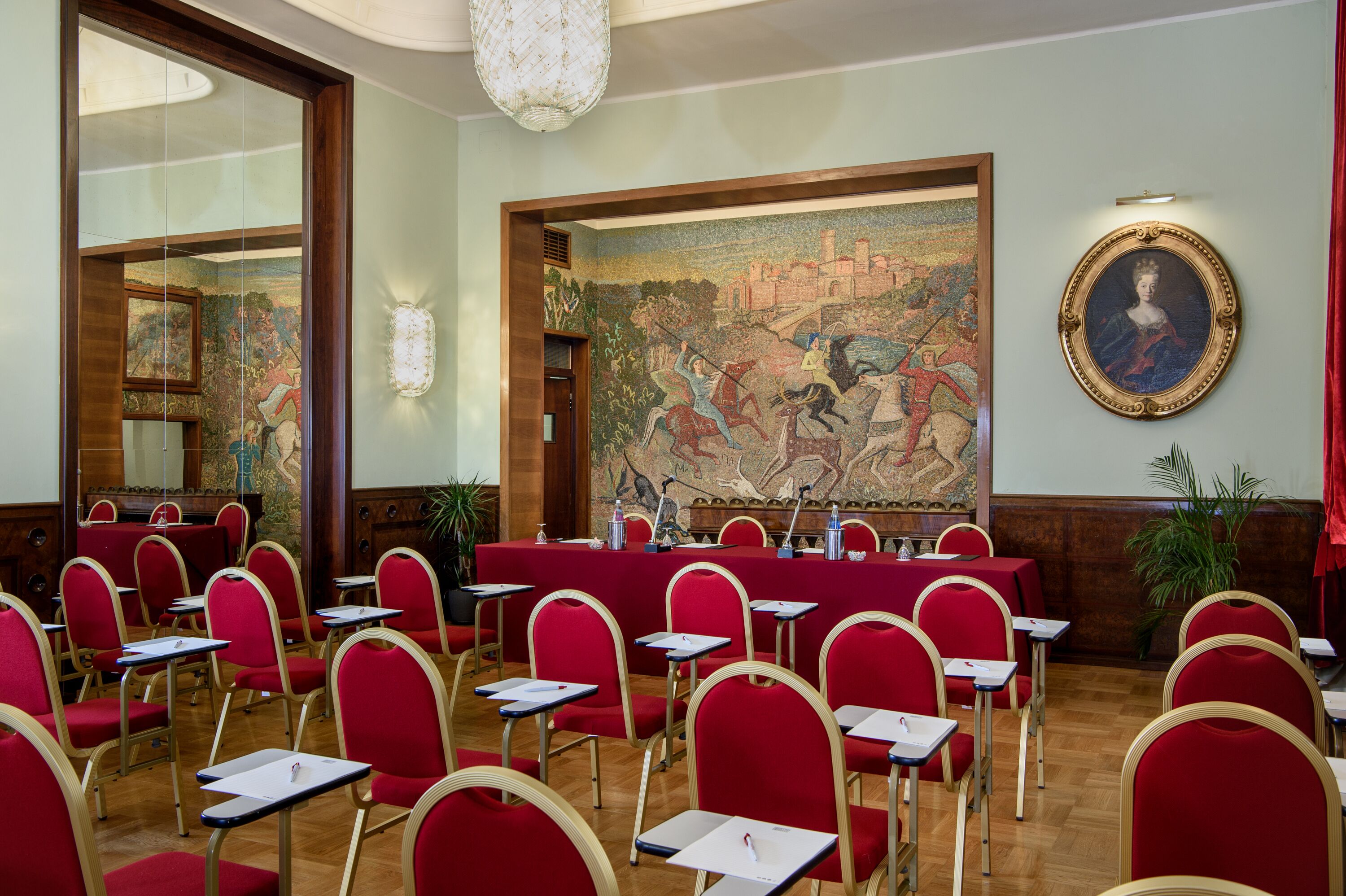 Uma sala de conferência com cadeiras e mesas vermelhas, um grande mural e um retrato emoldurado na parede.