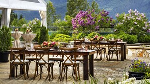 Uma área de refeições ao ar livre com mesas e cadeiras, rodeada por vasos de plantas e flores, com vista para a montanha ao fundo.