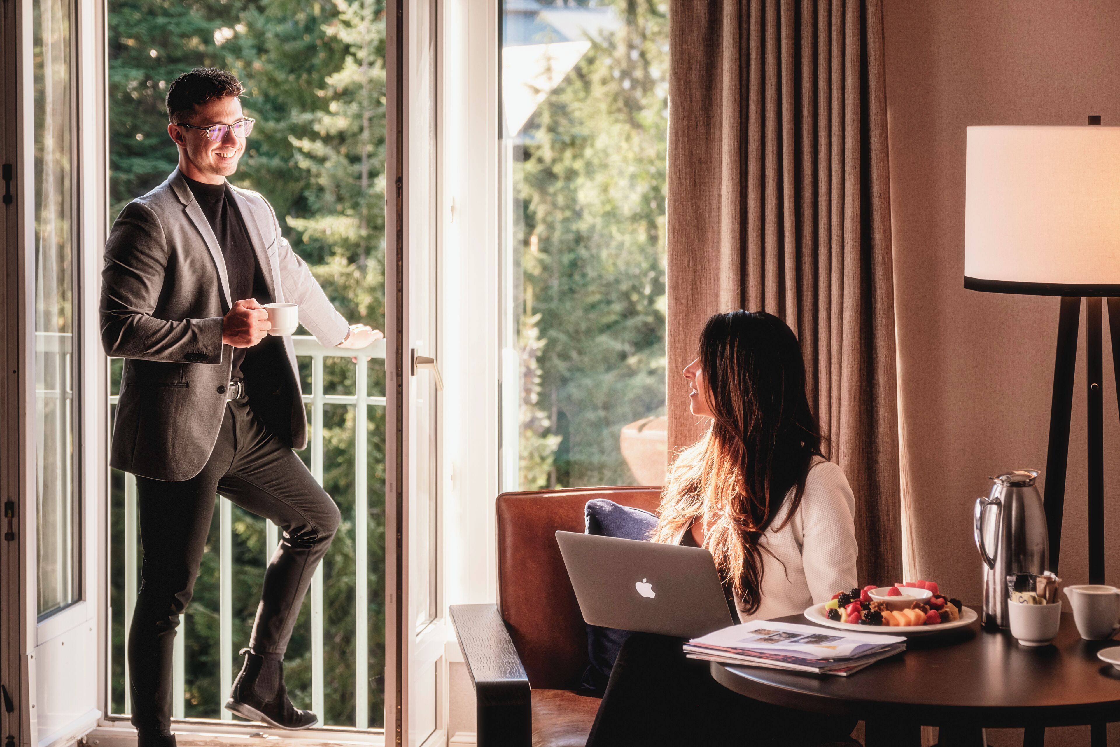 Um homem de terno ao lado de uma janela, uma mulher usando um laptop numa mesa com uma cafeteira e fruta.