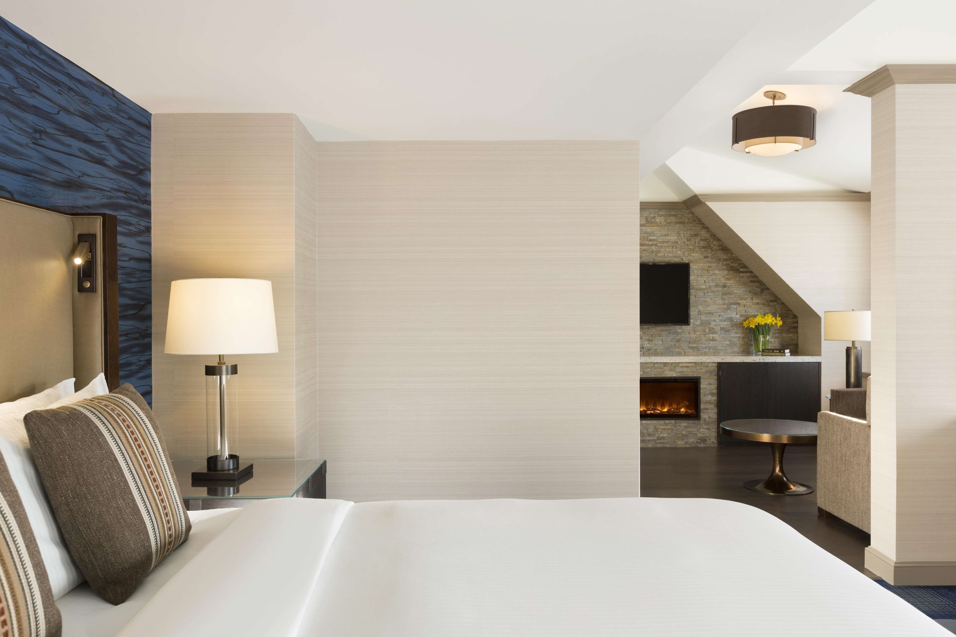 Quarto de hotel moderno com uma cama grande, escrivaninha com luminária, lareira e vista para o exterior.