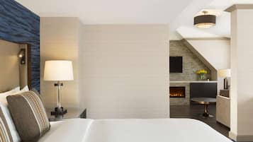 Quarto de hotel moderno com uma cama grande, escrivaninha com luminária, lareira e vista para o exterior.