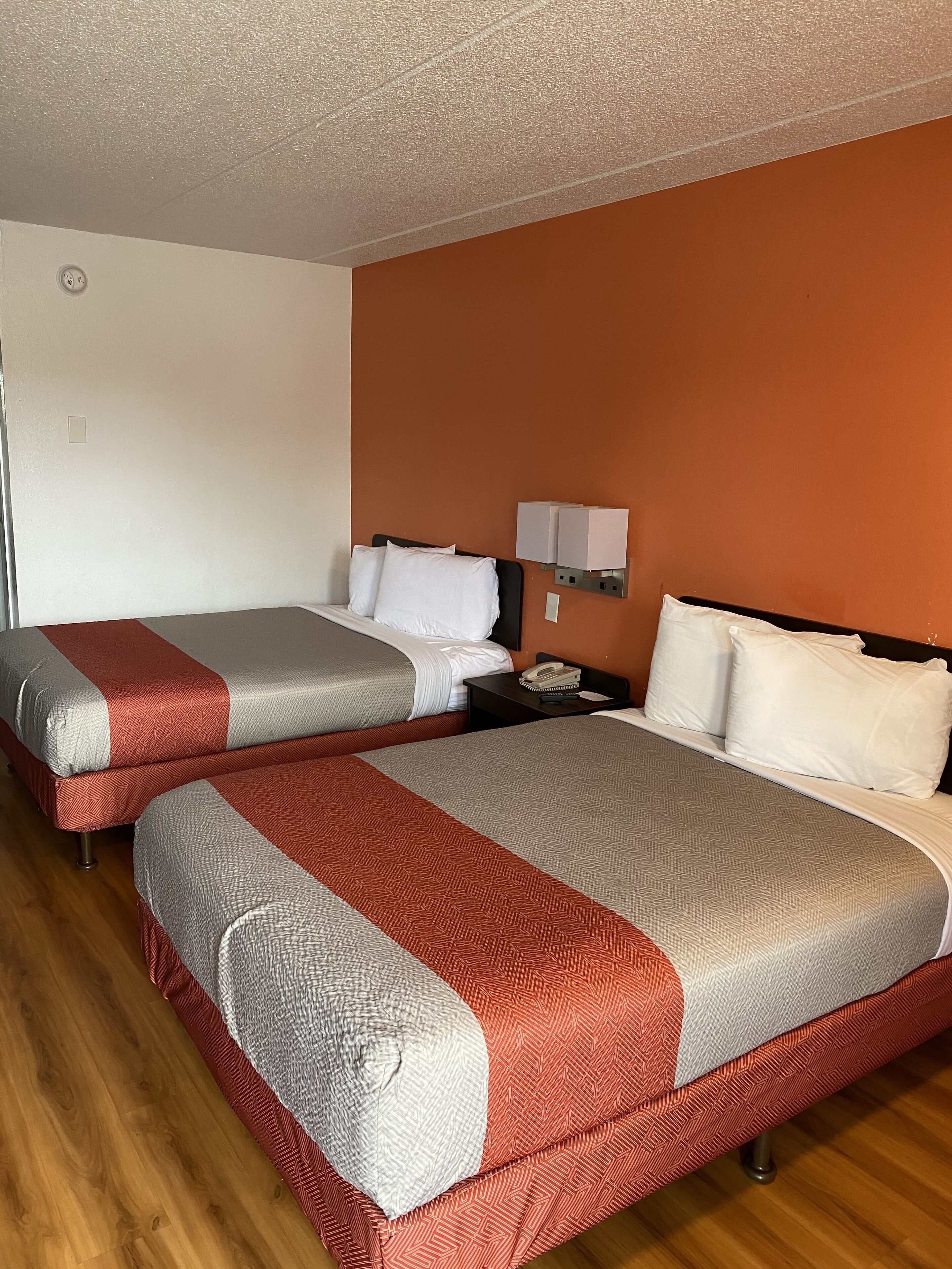 Duas camas de casal com roupa de cama estampada, piso de madeira, telefone fixado na parede e interruptor de luz.