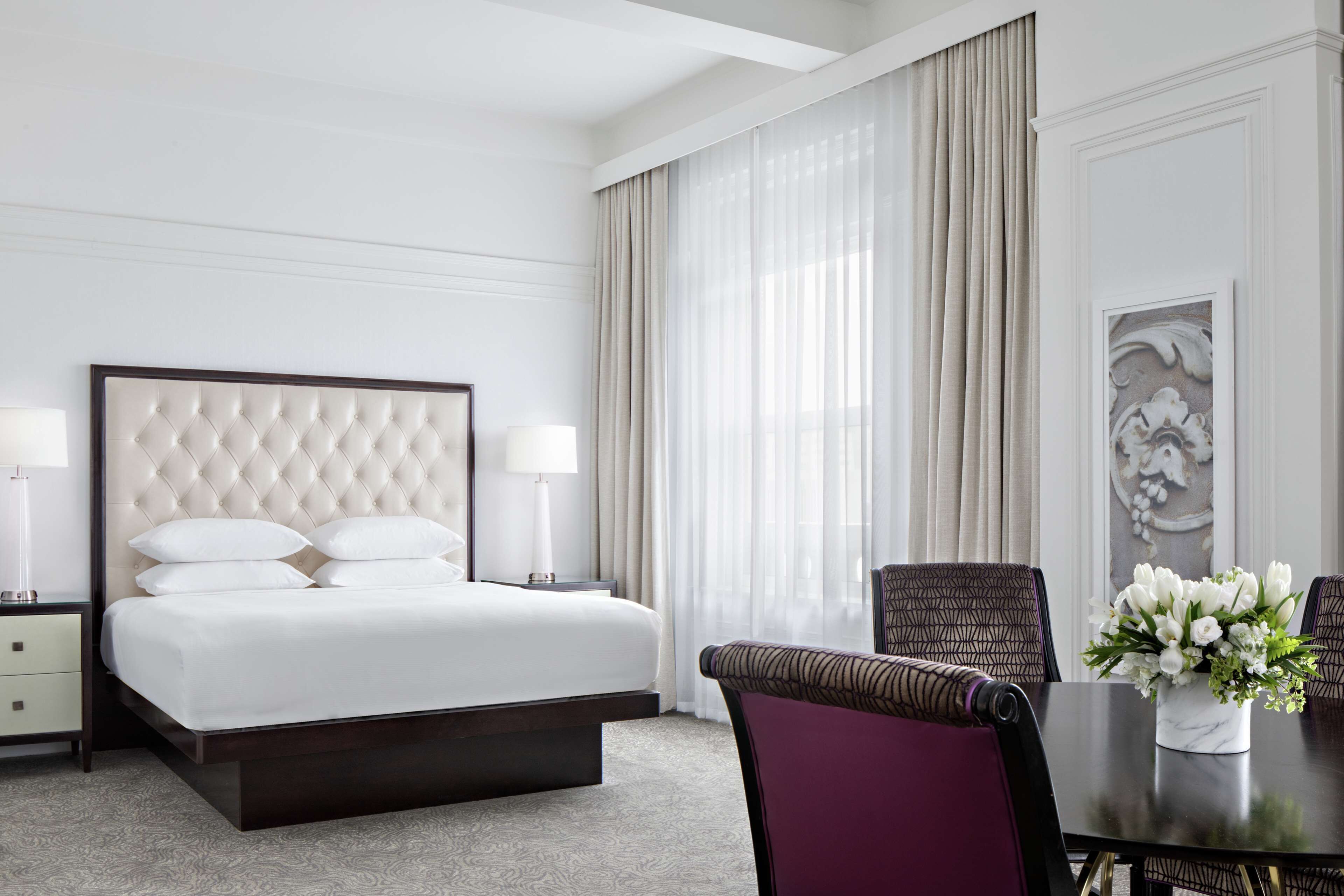 Quarto de hotel com uma cama grande, uma mesa de jantar com cadeiras e um vaso de flores.