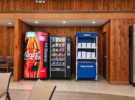 Uma área de distribuição automática com uma máquina de Coca-Cola, uma máquina de lanches e um carrinho de devolução de toalhas em um ambiente com acabamento em madeira.