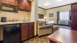 Quarto de hotel compacto com cozinha compacta, uma cama com cabeceira, um sofá, uma mesa pequena e uma janela com cortinas.