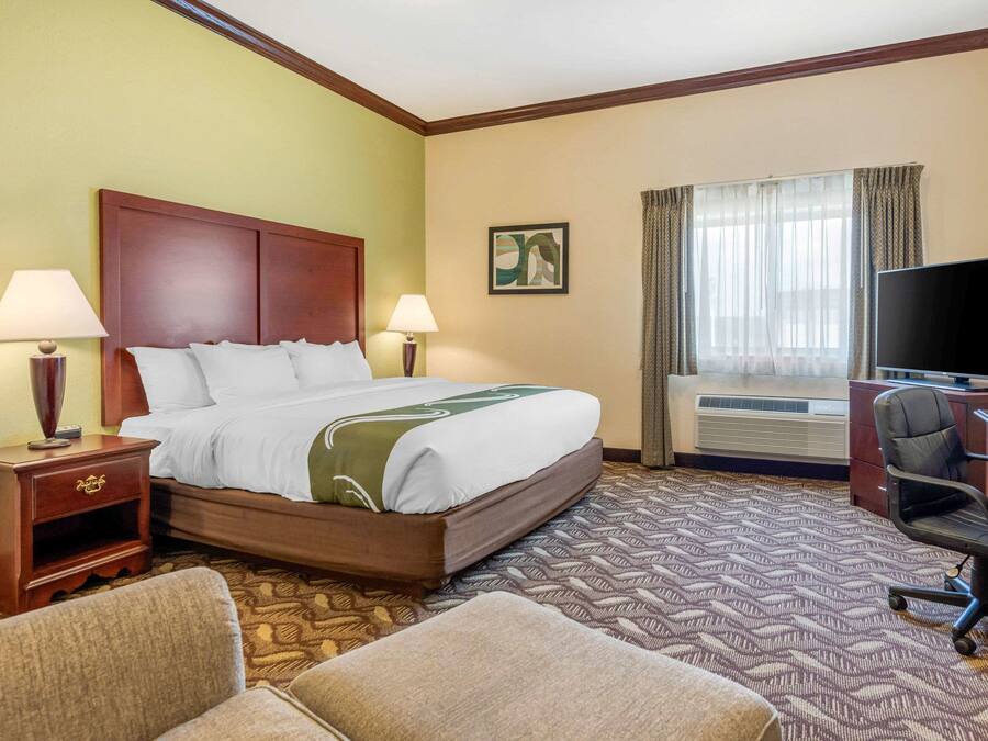 Quarto de hotel com cama, duas mesas de cabeceira com luminárias, escrivaninha com cadeira, televisão e ar condicionado.