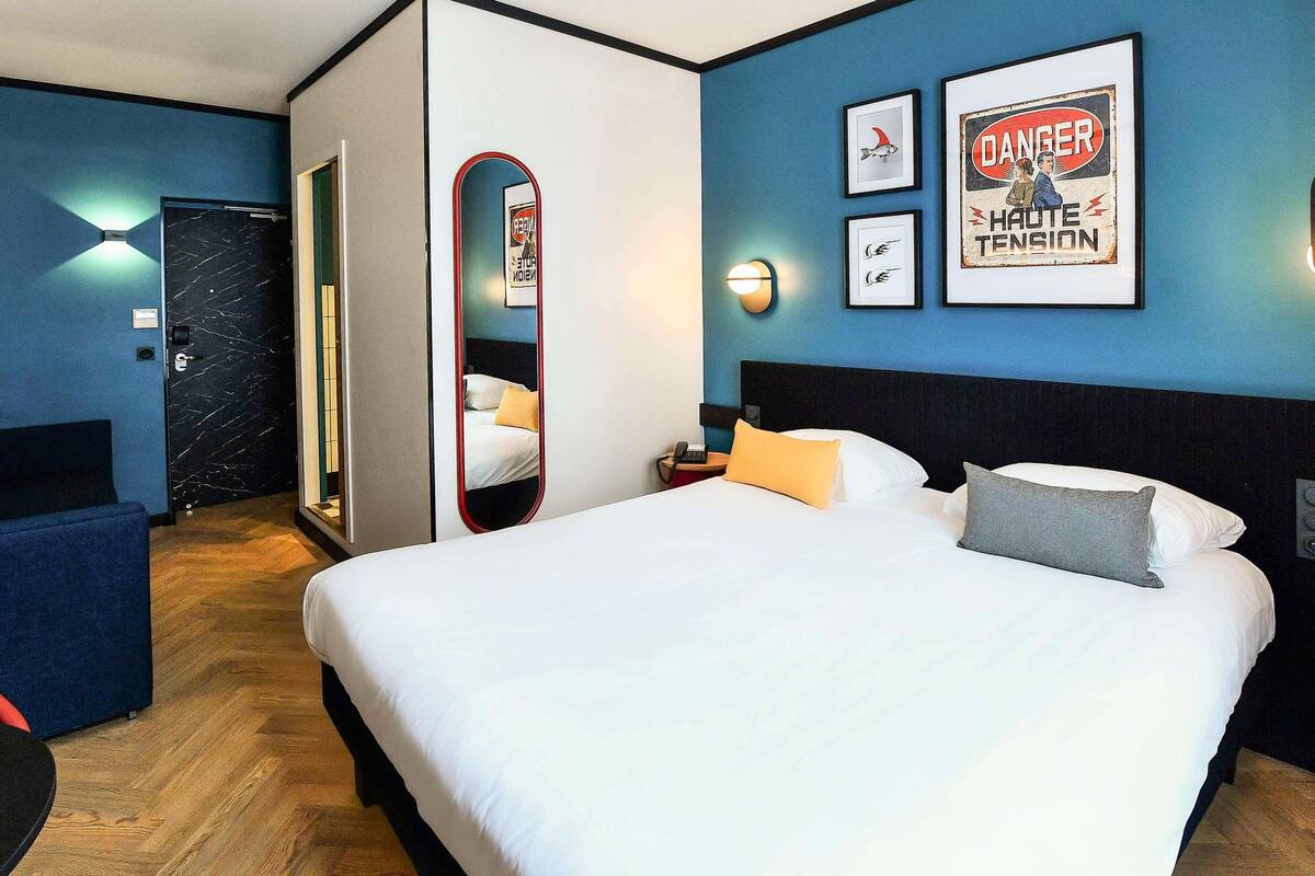 Quarto de hotel com uma cama grande, mesa de cabeceira com abajur, espelho e quadros na parede.