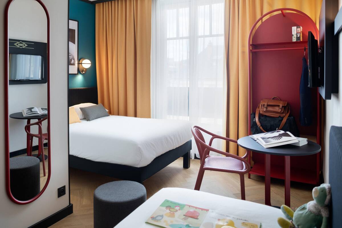 Quarto de hotel com cama, escrivaninha, cadeira, mesinha, espelho e guarda-roupa.