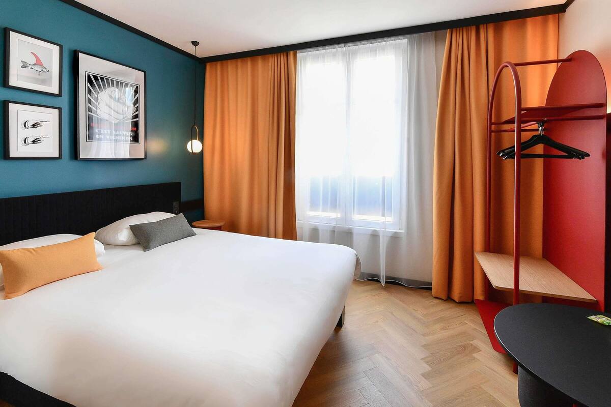 Quarto de hotel com cama, escrivaninha, cadeira e janela com cortinas.
