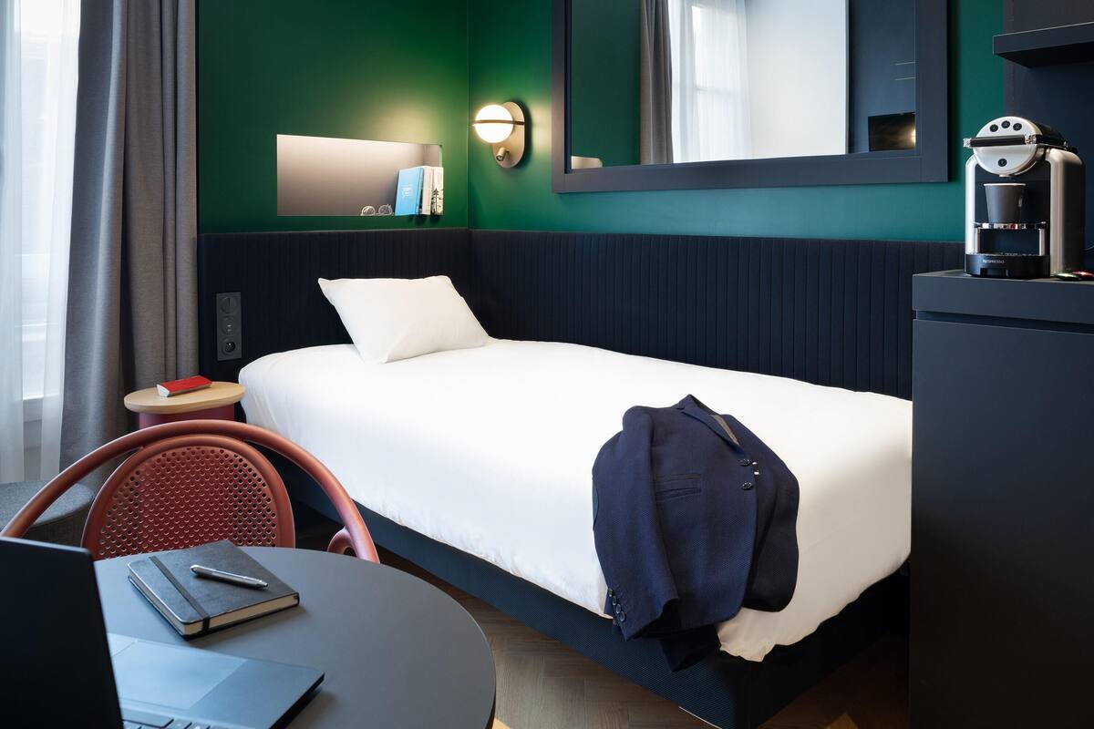 Quarto de hotel com cama, escrivaninha com laptop, cadeira, cafeteira e espelho.