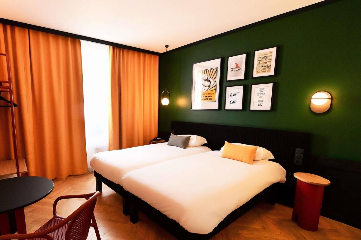 Quarto de hotel com duas camas, uma mesa, uma cadeira, um cesto de lixo e uma obra de arte na parede.
