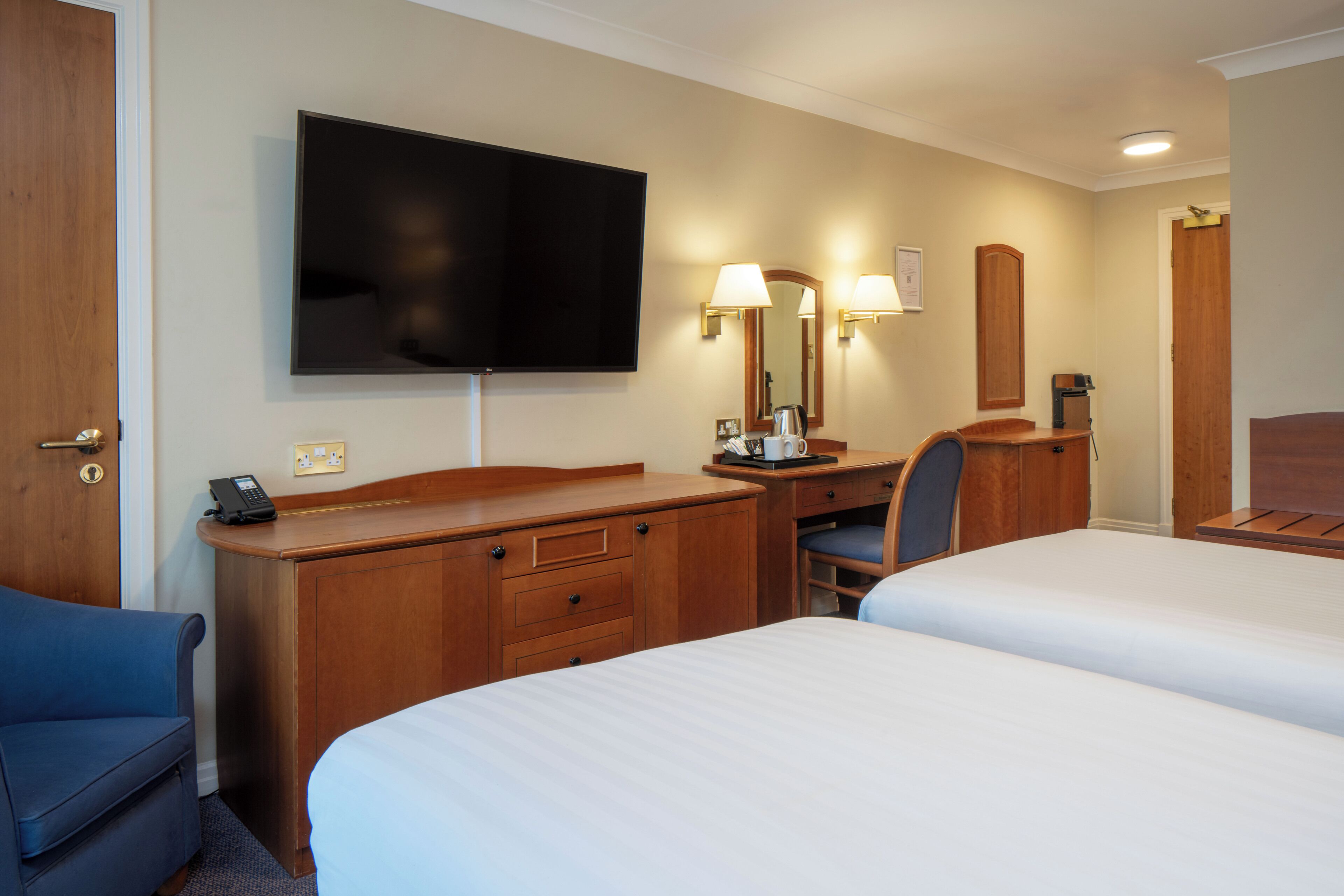 Quarto de hotel com televisão de tela plana, uma escrivaninha com cadeira, mesa de cabeceira e duas camas.