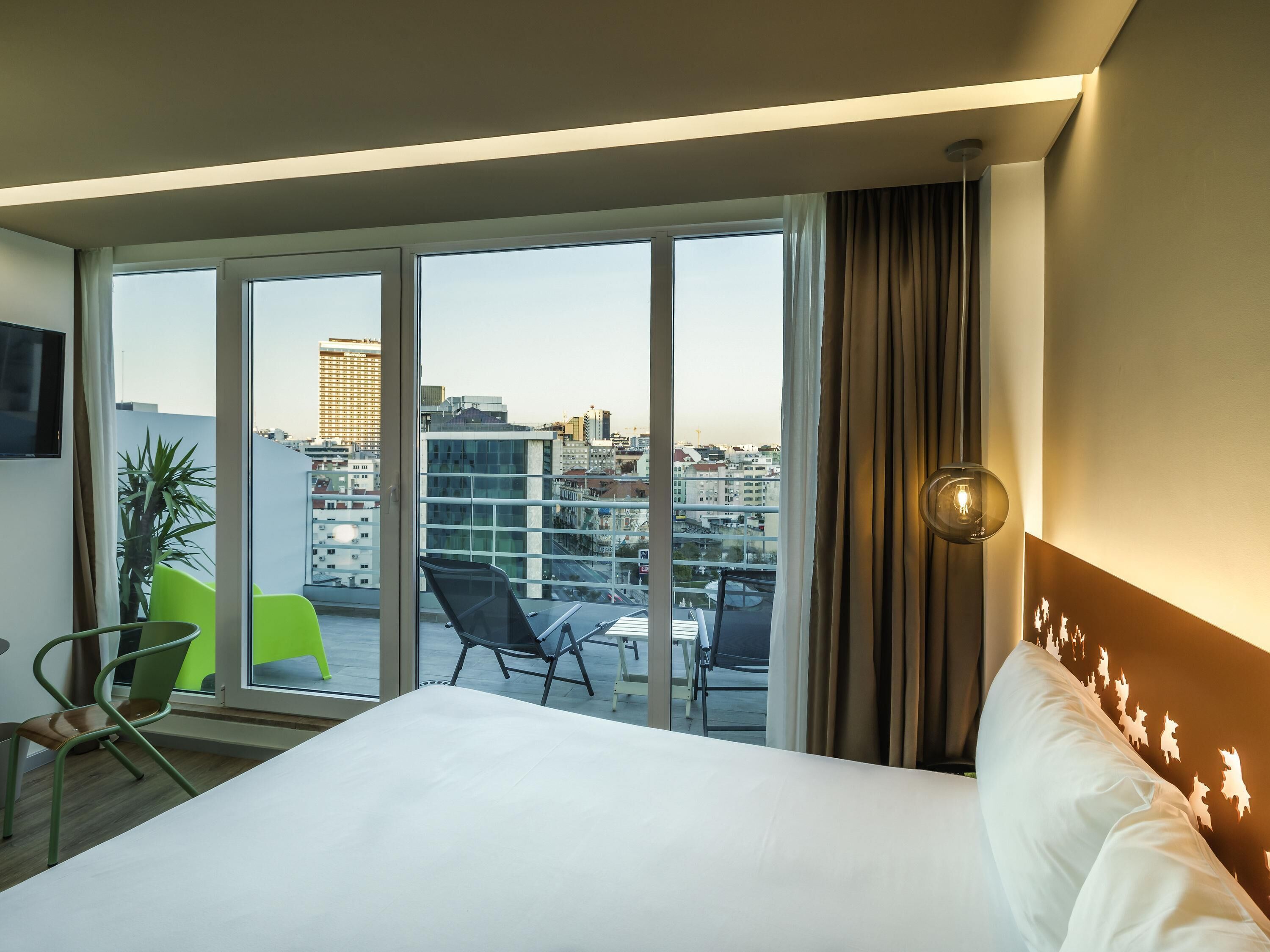 Quarto de hotel com uma cama grande, vista para a cidade, televisão na parede e uma planta.