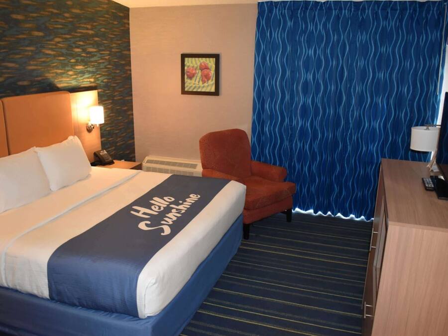 Quarto de hotel com cama, uma cadeira, escrivaninha, abajur e um quadro na parede.