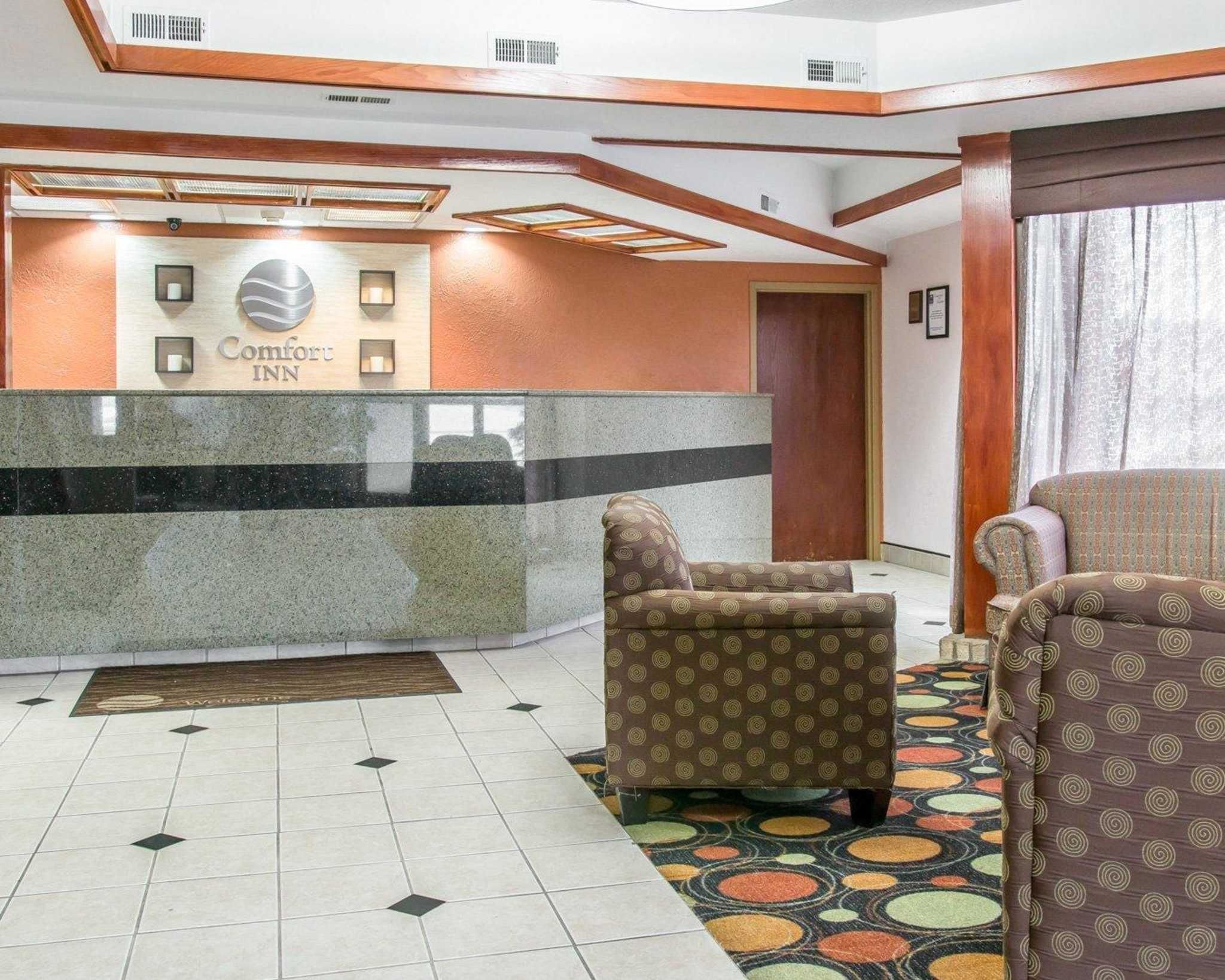 Uma área de recepção de hotel, com balcão de atendimento, duas poltronas estampadas, tapete colorido e teto de madeira.