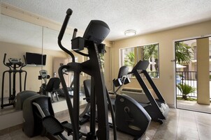 Uma academia com diversos equipamentos de exercício, uma televisão e vista para o exterior através de uma porta de vidro.