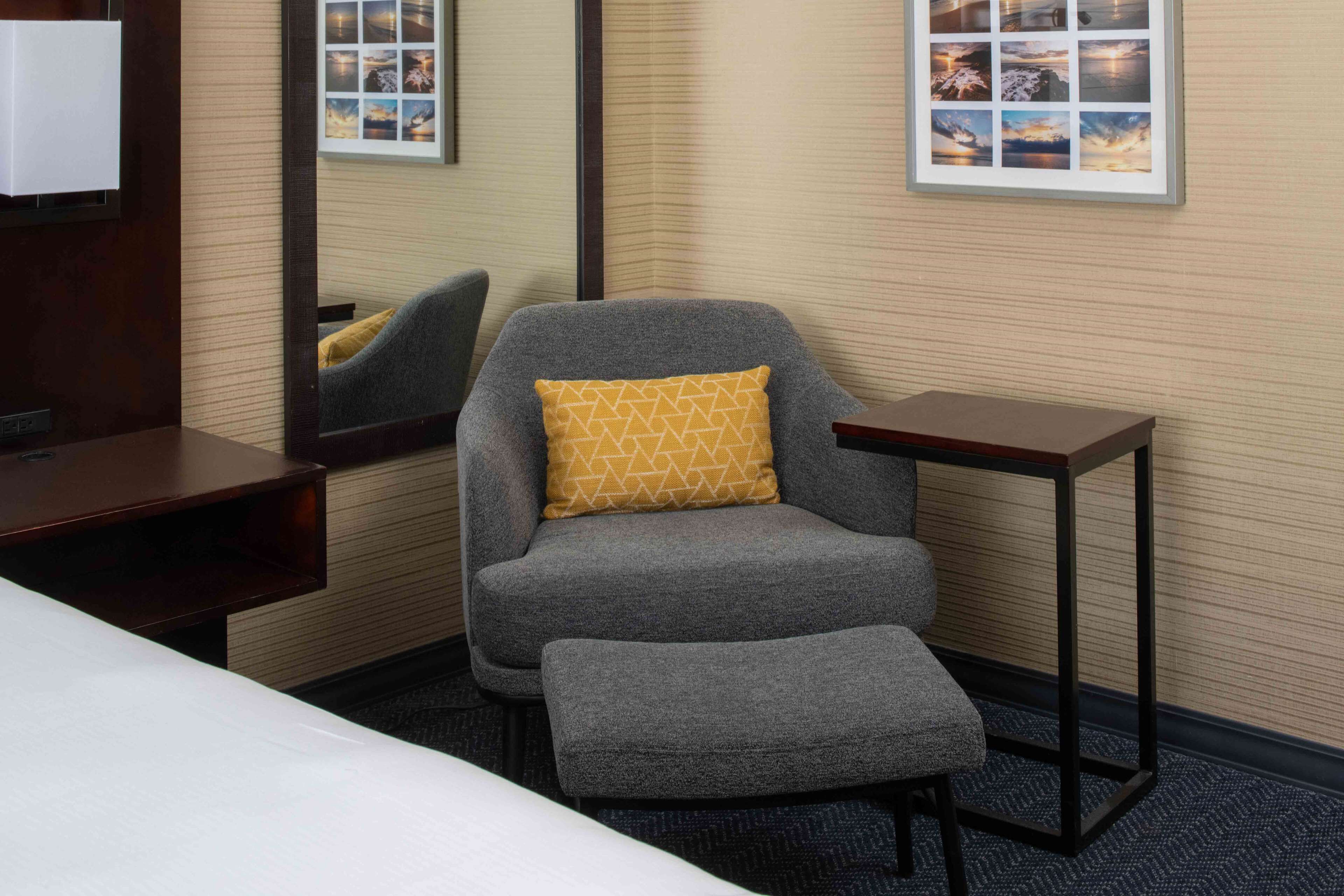Quarto de hotel com uma poltrona cinzenta, uma mesa pequena, uma mesinha de cabeceira e uma cama.