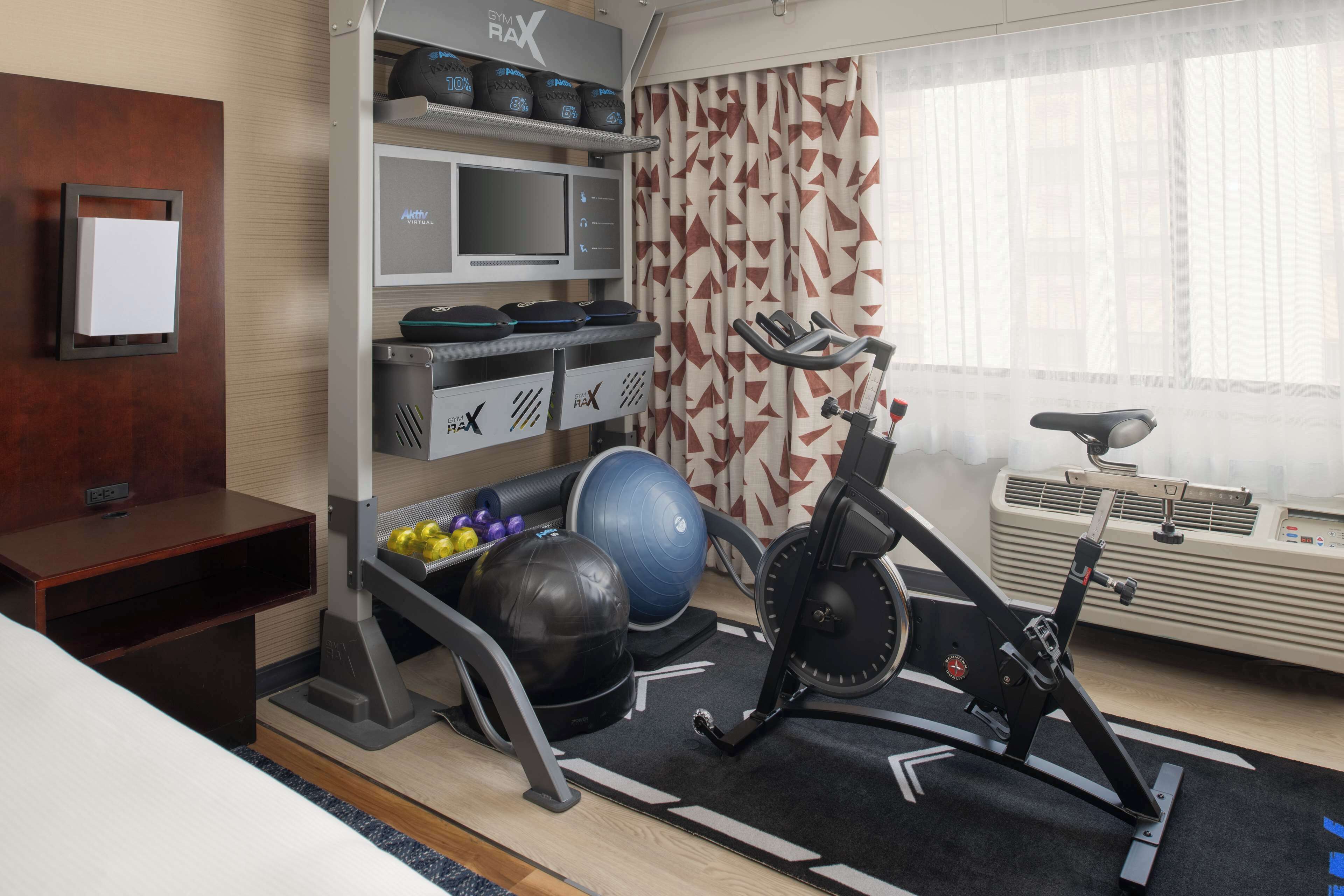 Uma academia com equipamentos de exercício, uma TV fixada na parede e uma cortina estampada.