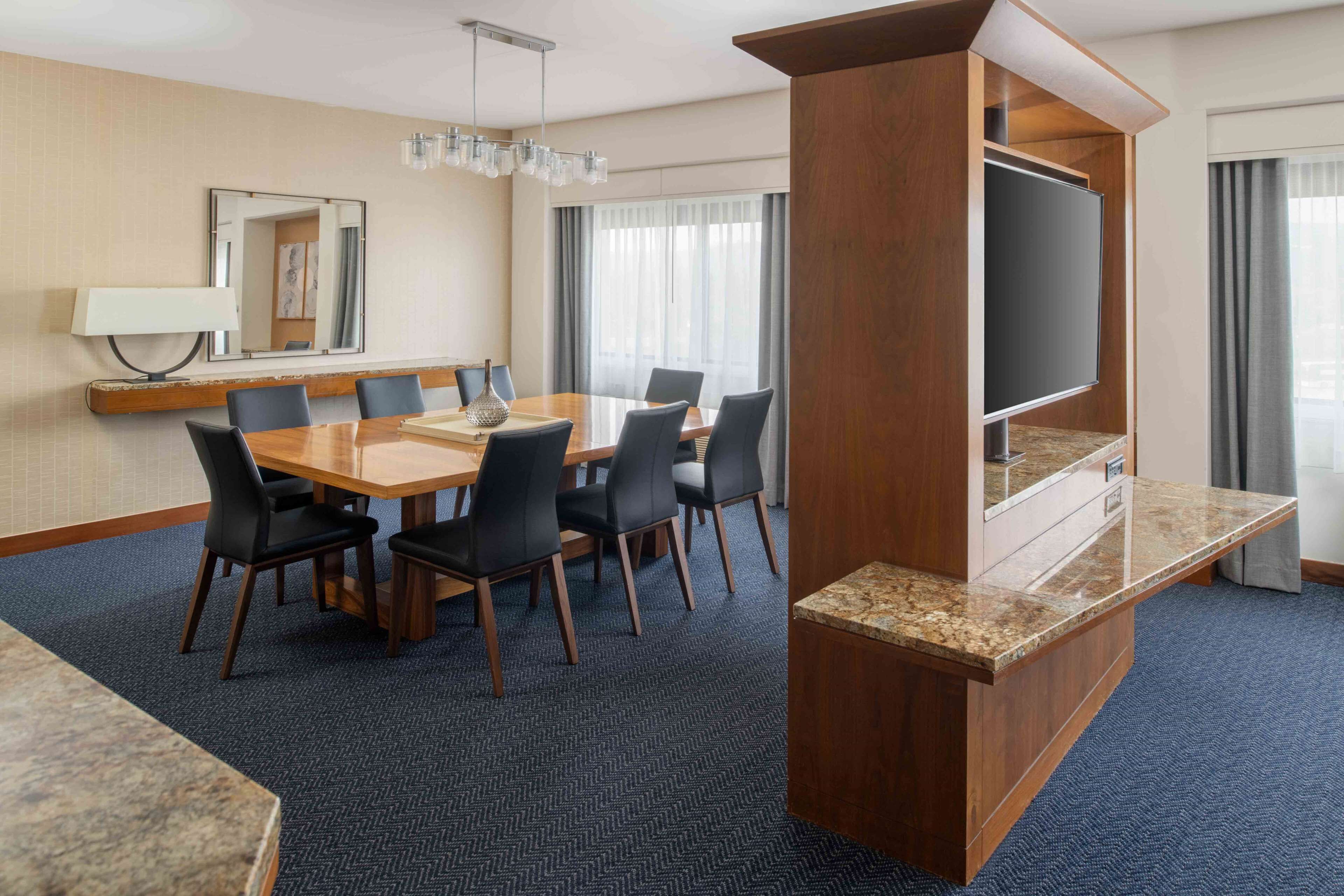 Quarto de hotel com área de refeições, apresentando uma mesa e cadeiras de madeira, televisão fixada em um móvel de madeira e uma janela grande com cortinas.