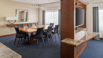 Quarto de hotel com área de refeições, apresentando uma mesa e cadeiras de madeira, televisão fixada em um móvel de madeira e uma janela grande com cortinas.