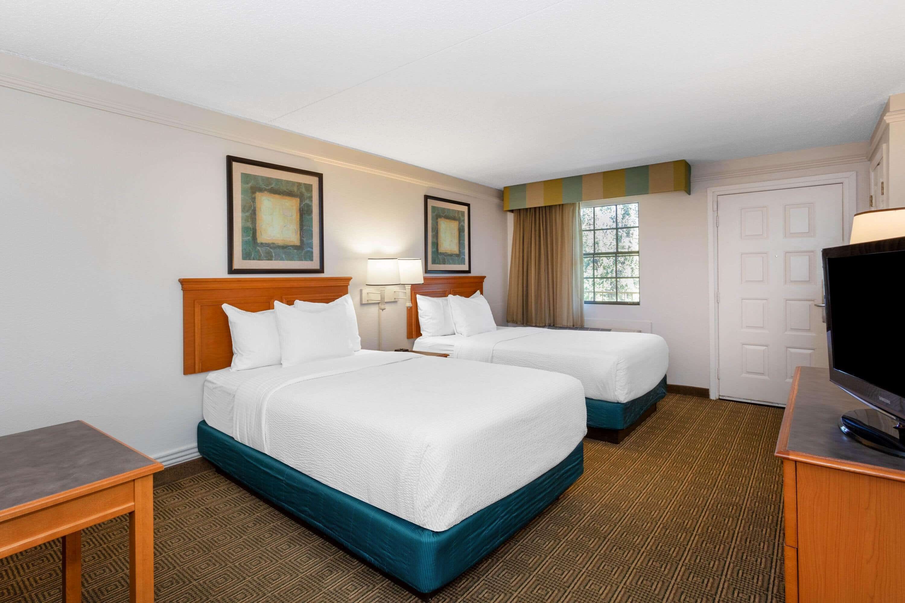 Quarto de hotel com duas camas, televisão, escrivaninha e janela com cortinas.