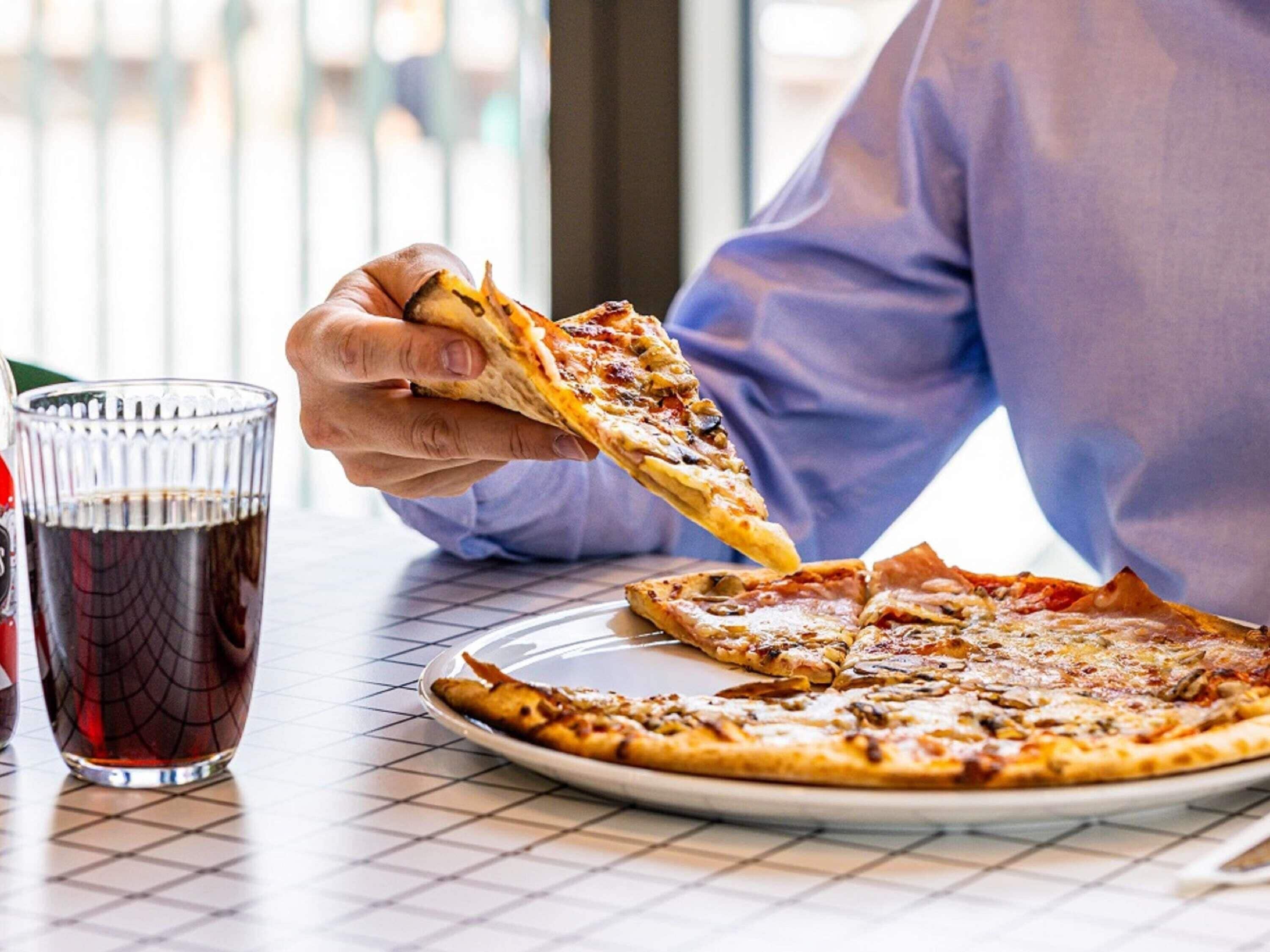 Uma pessoa segurando uma fatia de pizza, com um copo de refrigerante e uma pizza sobre a mesa.