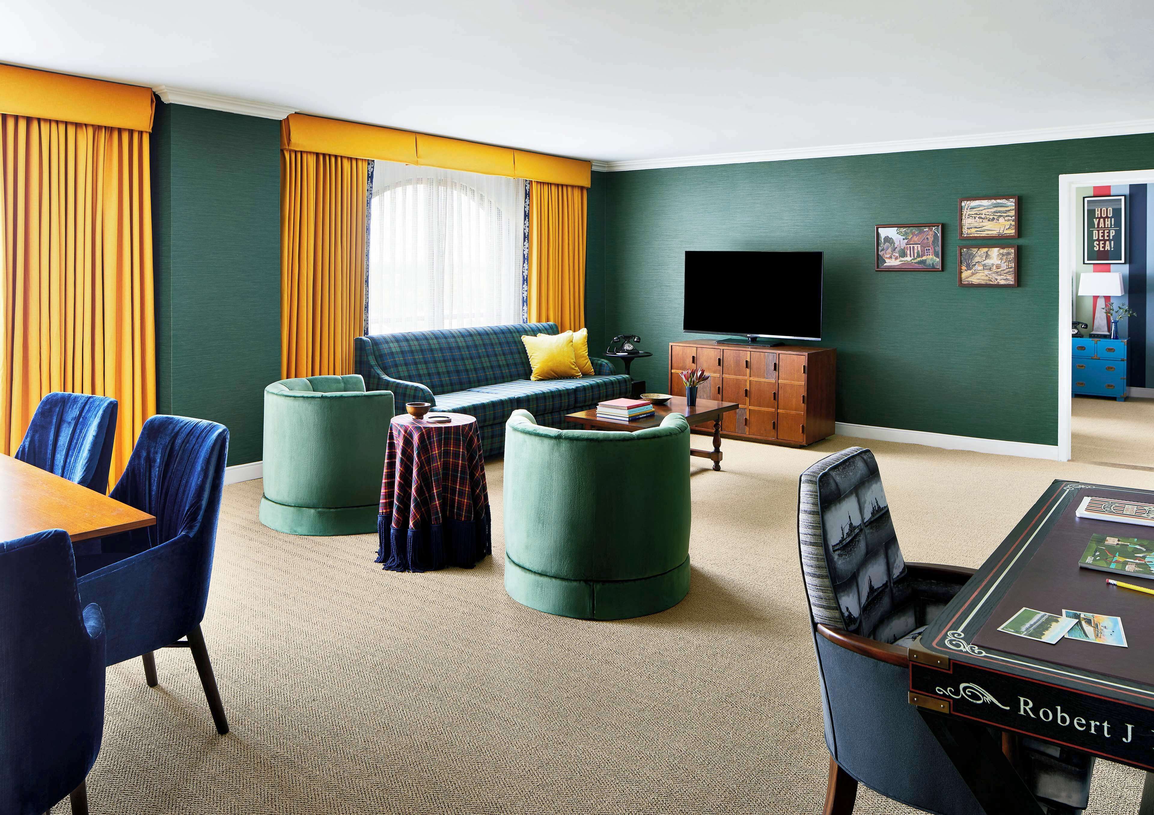 Uma sala de estar com um sofá verde, poltronas azuis, uma mesa de centro e uma televisão.