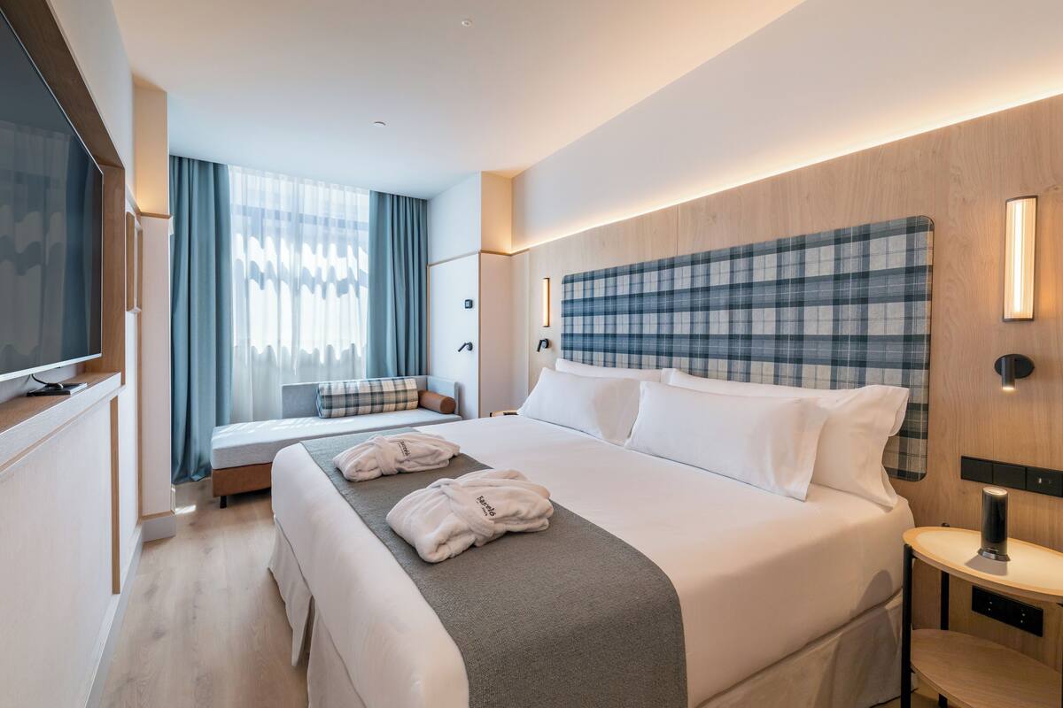 Quarto de hotel com uma cama grande, televisão, área de estar com sofá e cortinas, e cabeceira com padrão xadrez.