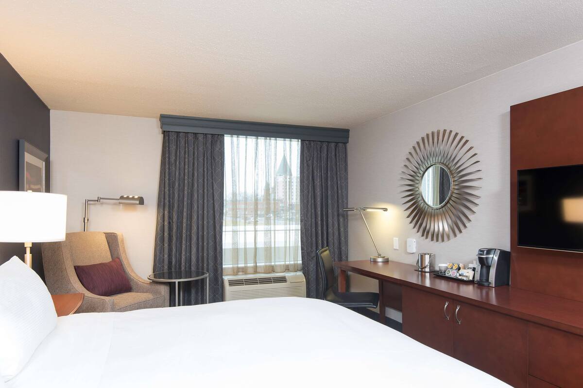 Quarto de hotel com cama, escrivaninha com cafeteira, televisão, espelho e cadeira com almofada.