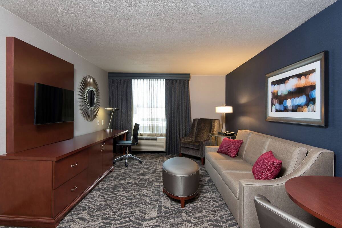 Quarto de hotel com televisão de tela plana, sofá, banquinho redondo, escrivaninha com cadeira e janela com cortinas.