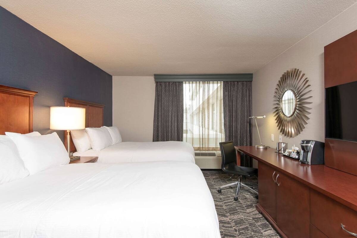 Quarto de hotel com duas camas, uma escrivaninha com cadeira, máquina de café, televisão e um espelho grande na parede.