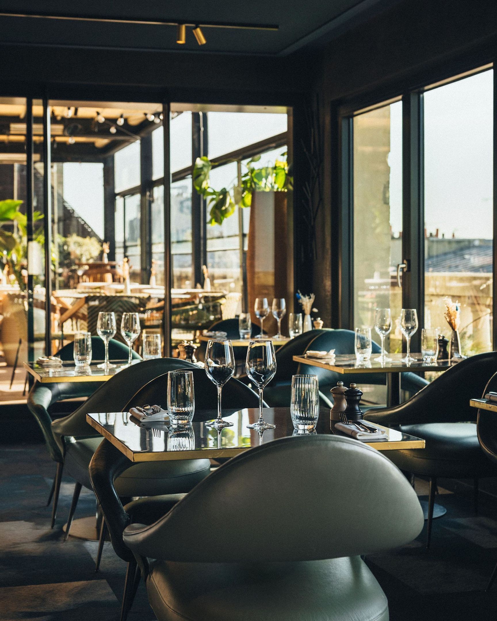 Interior de restaurante com mesas preparadas para refeições, janelas amplas e plantas.