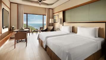 Quarto de hotel com duas camas, varanda com vista para o mar, área de refeições e ventoinha no teto.