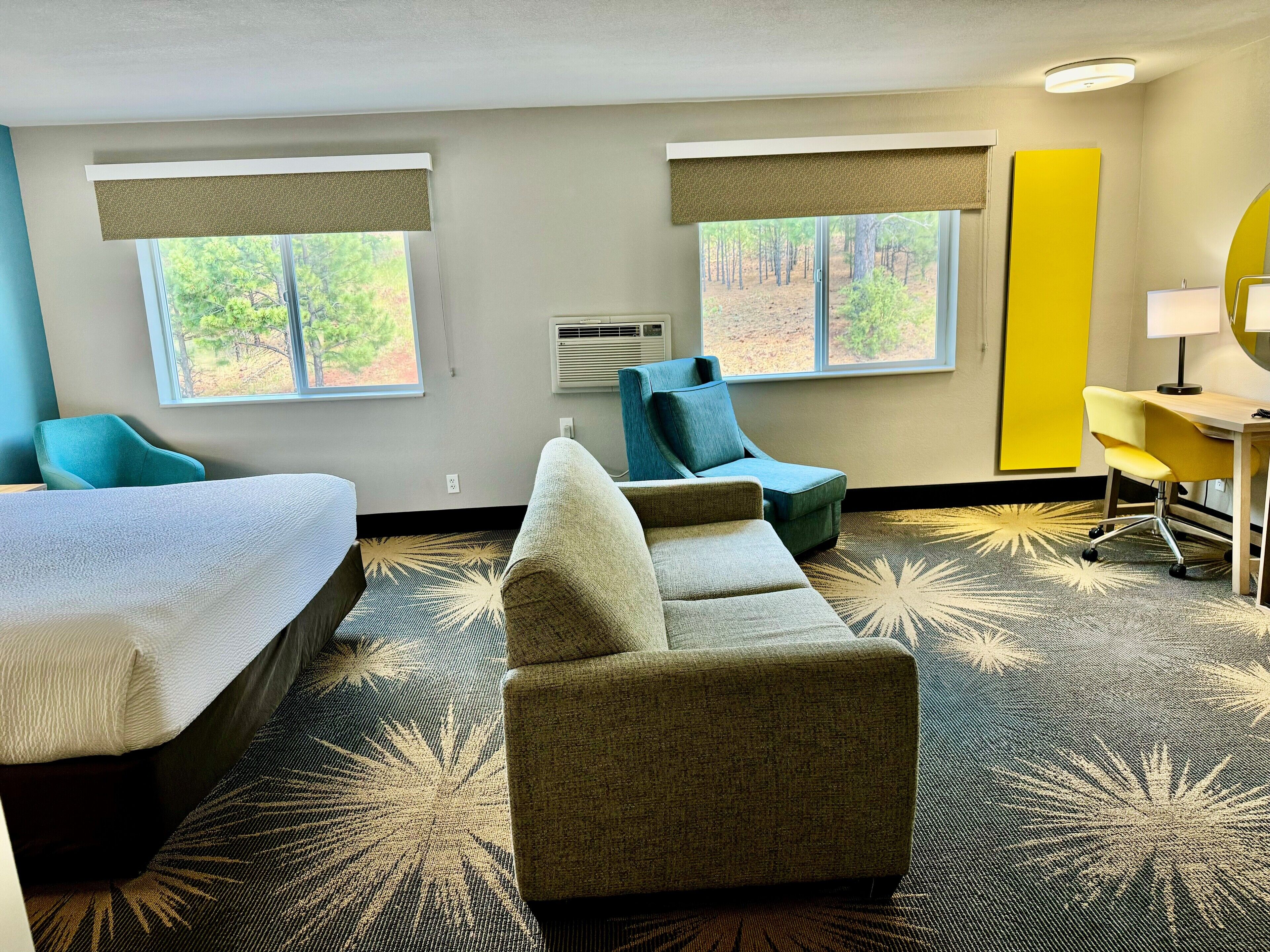 Quarto de hotel com uma cama, duas poltronas, uma secretária com abajur e vista para árvores pelas janelas.