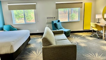 Quarto de hotel com uma cama, duas poltronas, uma secretária com abajur e vista para árvores pelas janelas.