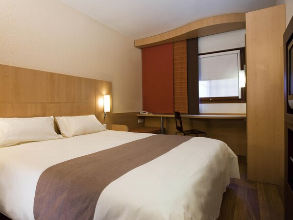Quarto de hotel com uma cama grande, uma escrivaninha, uma cadeira e uma televisão.