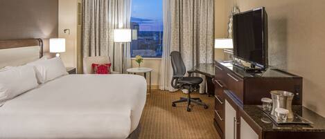 Quarto de hotel com uma cama grande, uma escrivaninha com televisão, uma cadeira e uma pequena mesa com abajur.
