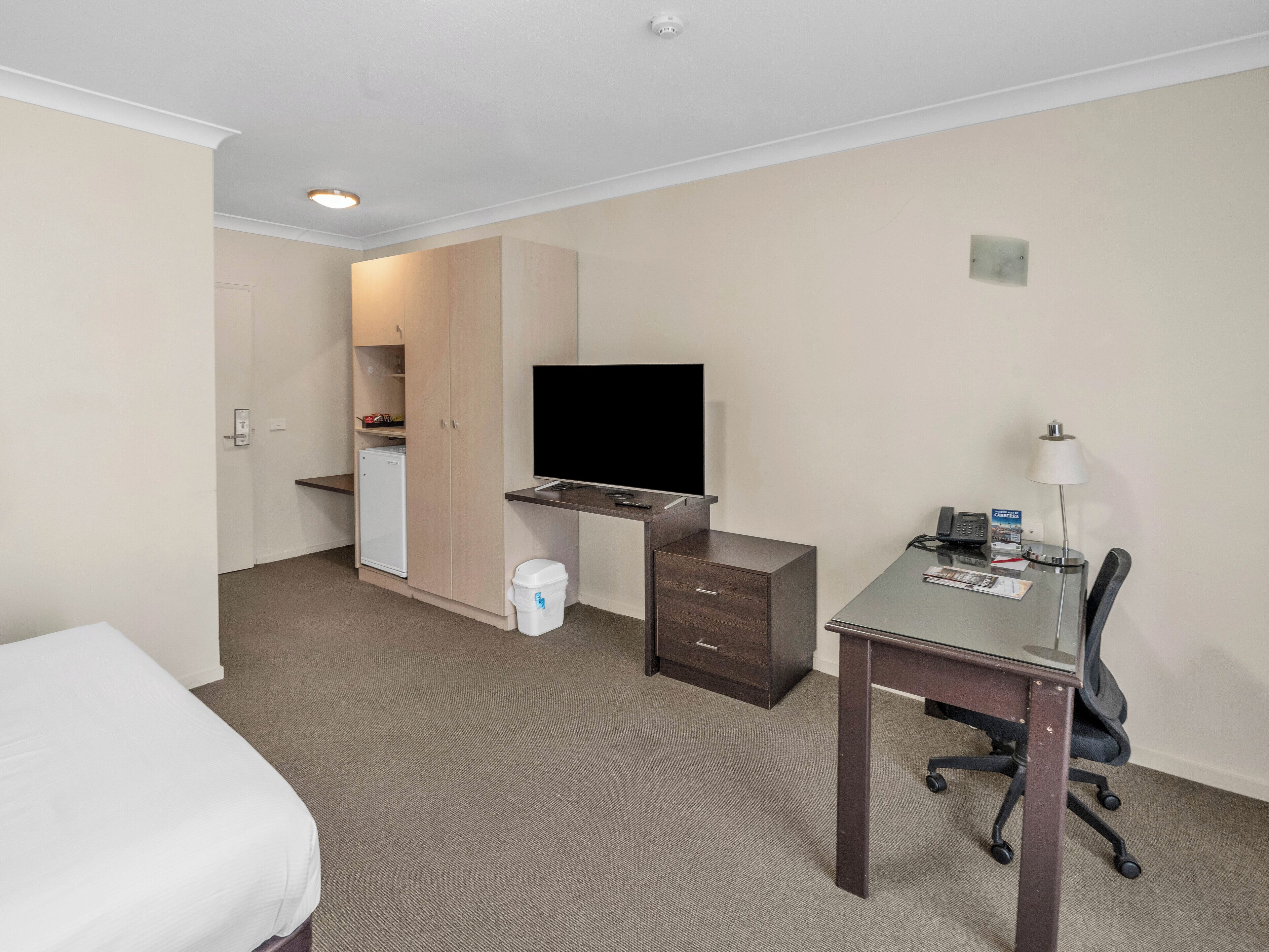 Quarto de hotel com cama, escrivaninha com cadeira, televisão, pequeno espaço de cozinha e lixeira.