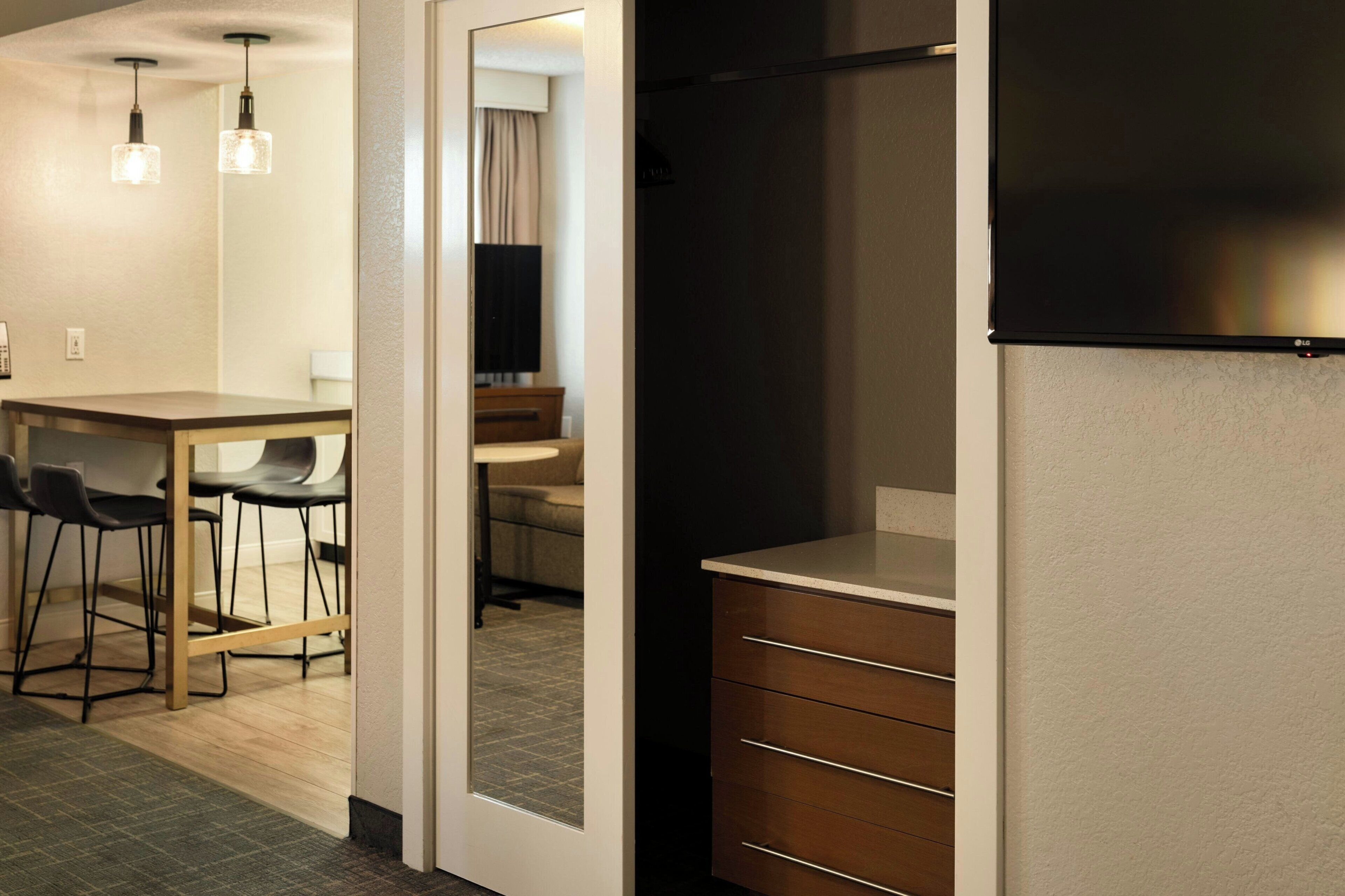 Quarto de hotel com cozinha compacta, área de refeições com cadeiras, espelho e guarda-roupa.