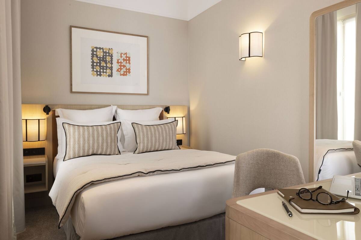 Quarto de hotel com uma cama, abajures, uma escrivaninha com um laptop, copos e um caderno.
