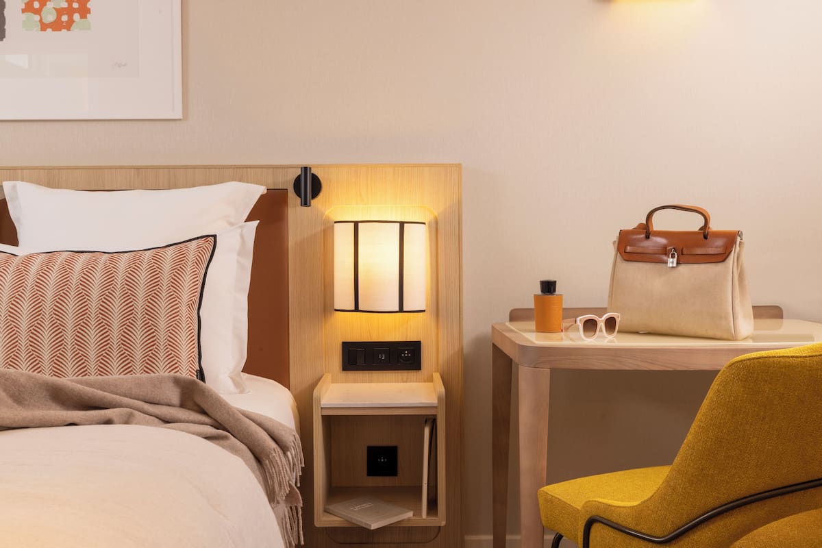 Quarto de hotel com cama, mesa de cabeceira com abajur, cadeira e uma bolsa sobre a mesa.